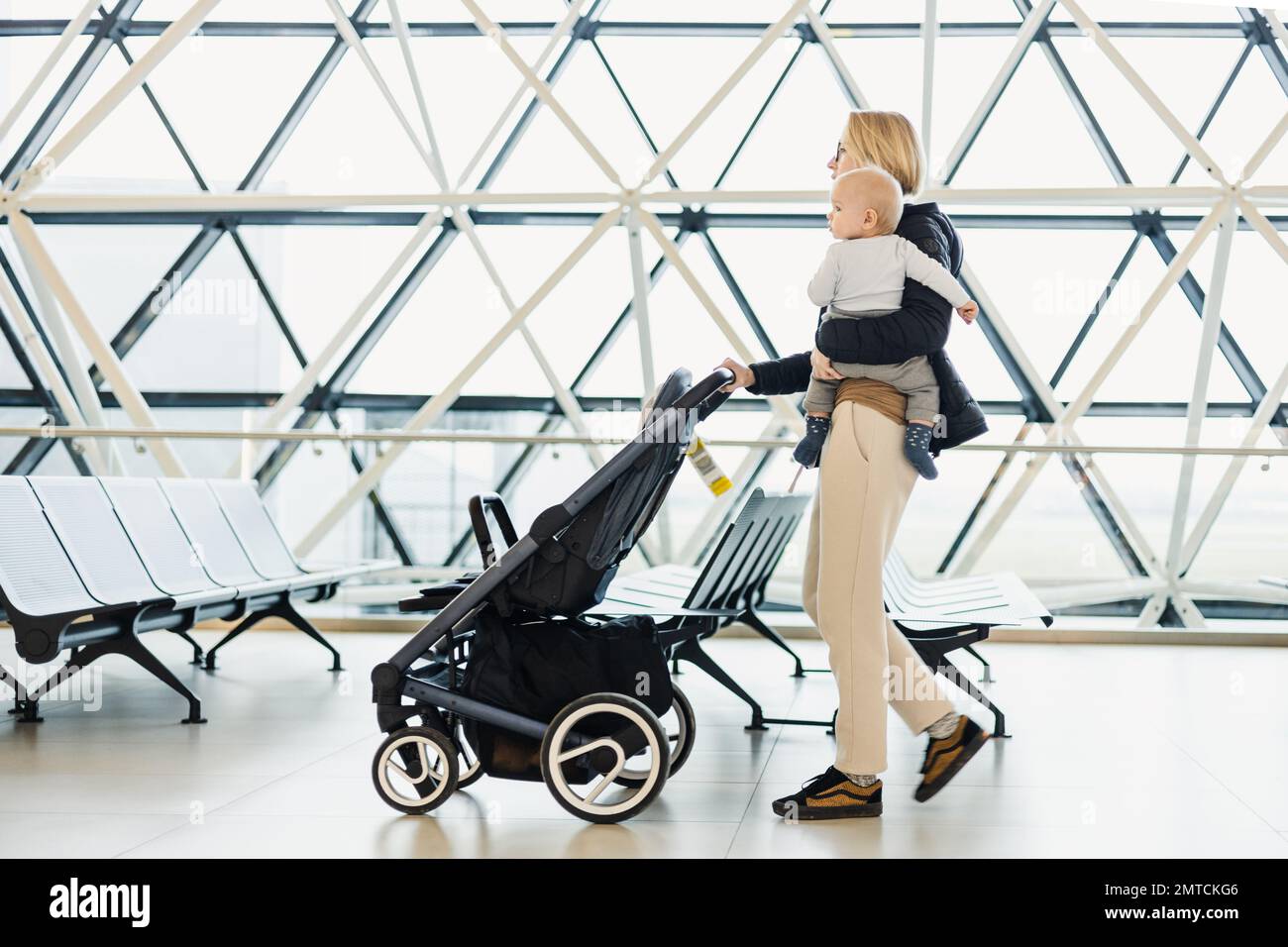 Mother carying his infant baby boy child, pushing stroller at airport ...