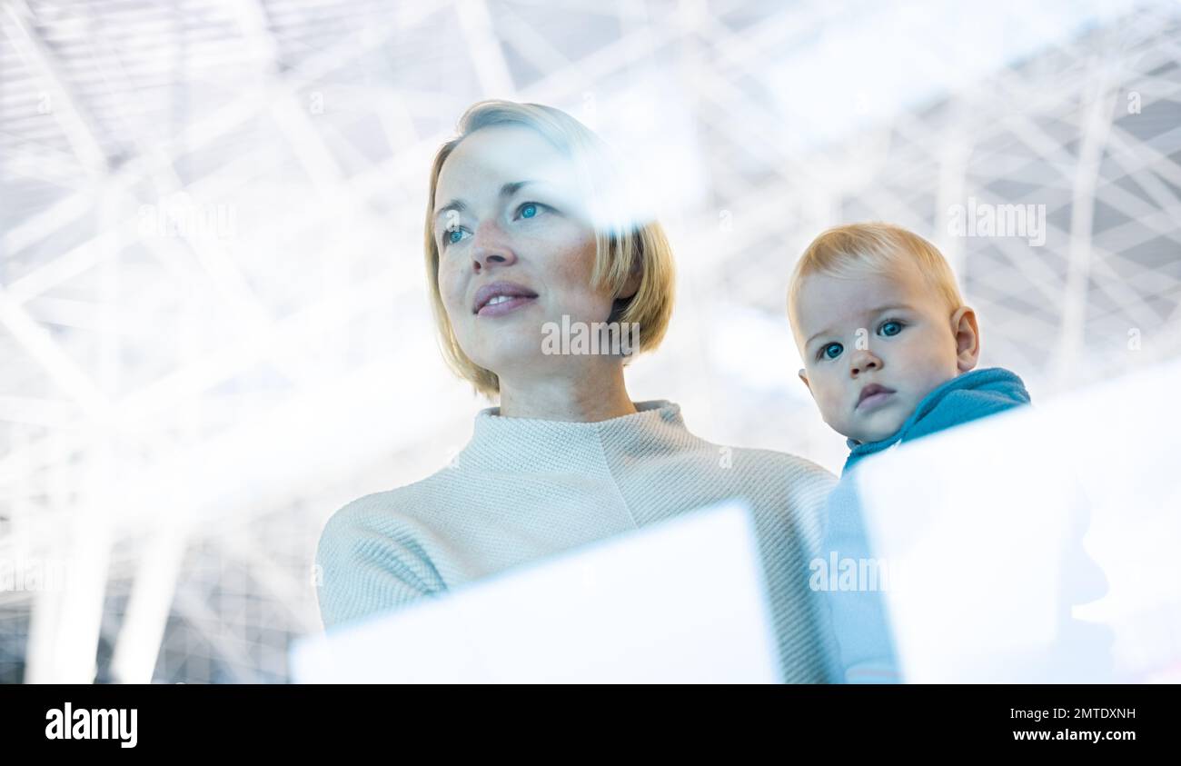 Thoughtful young mother looking trough window holding his infant baby ...