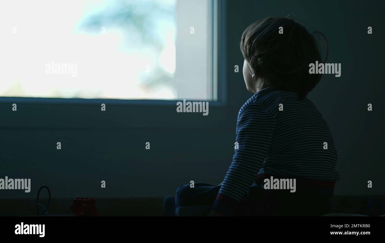 One lonely small boy sitting in the dark staring at window. Depressed ...