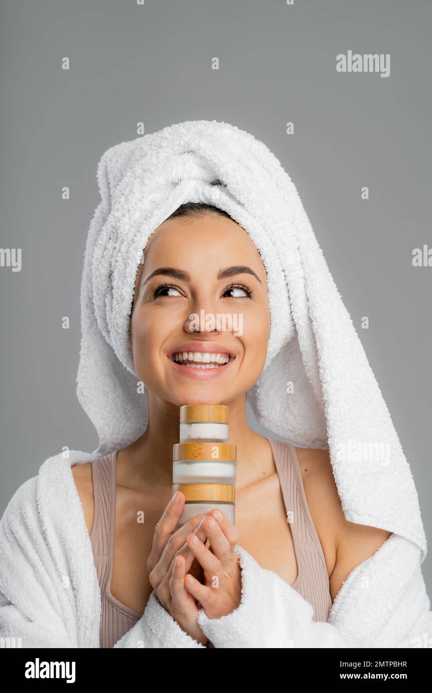 Positive Young Woman In Towel On Head Holding Cosmetic Creams Isolated