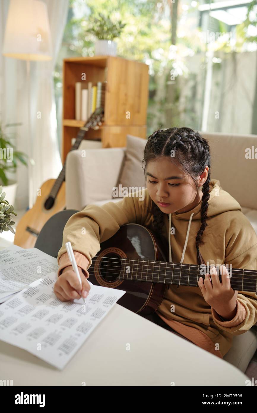 Concentrated teenage girl writing down music notes when playing guitar ...