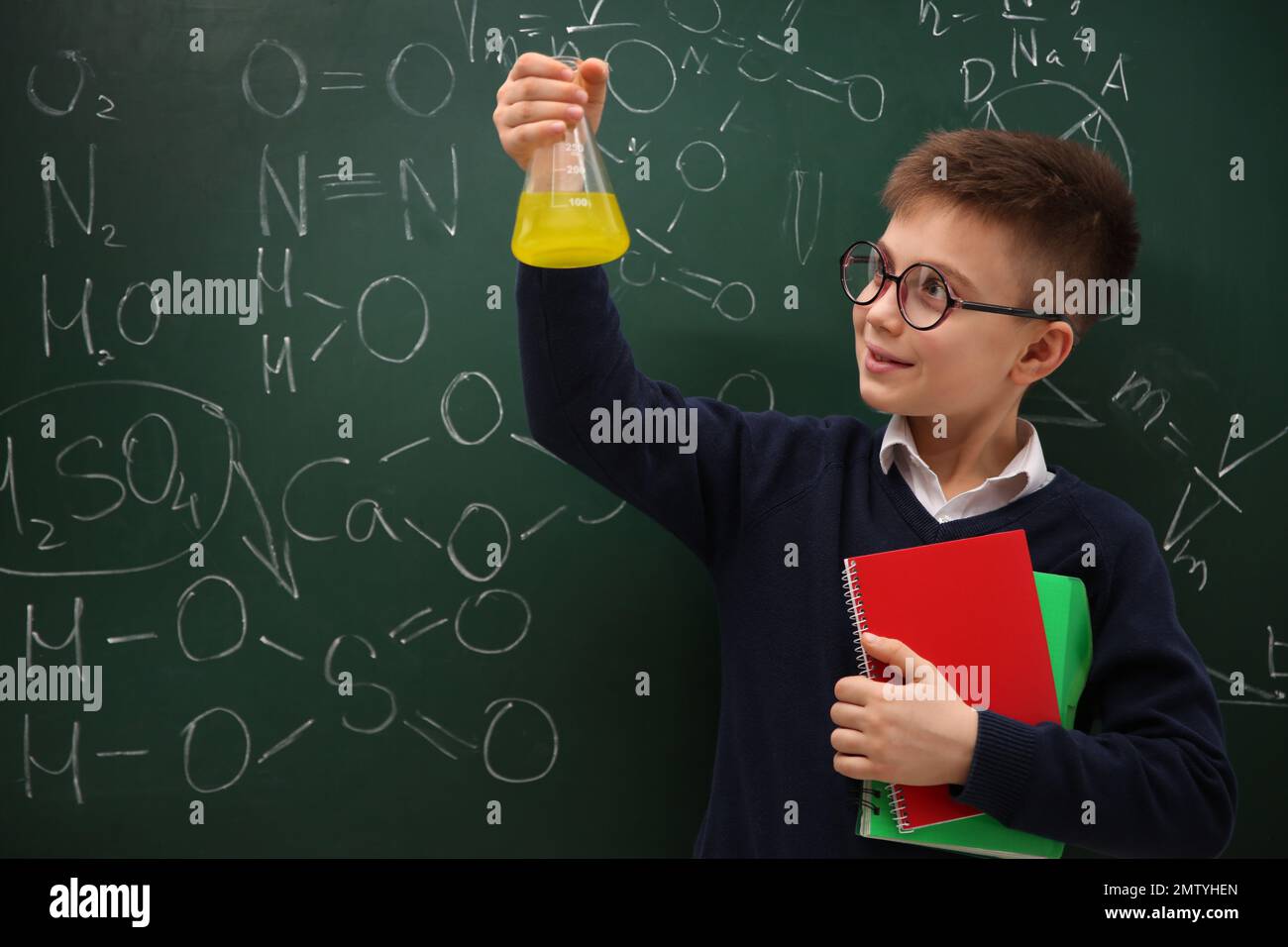 Schoolboy holding flask and notebooks near chalkboard with chemical ...