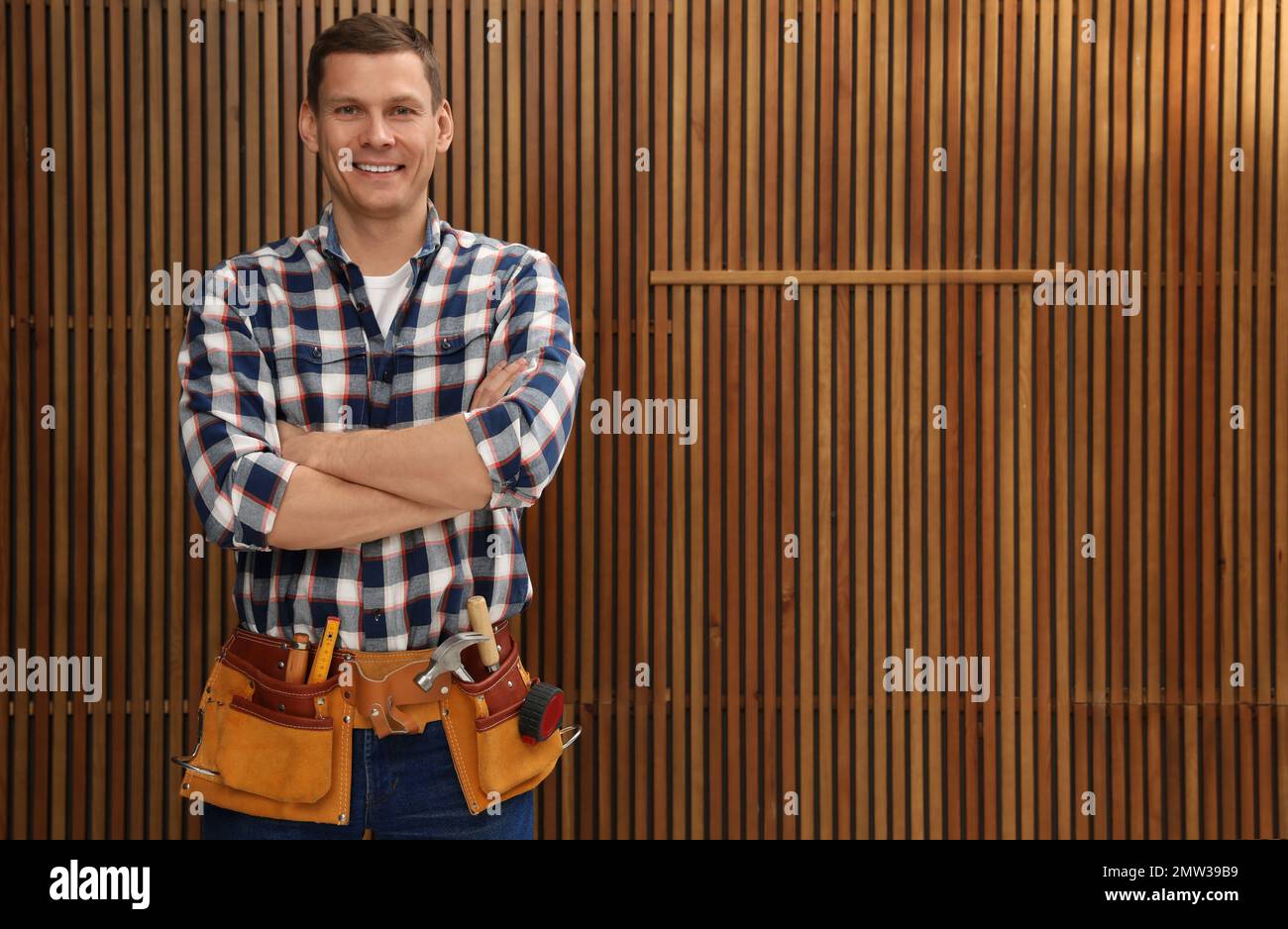 Handsome carpenter with tool belt near wooden wall. Space for text ...