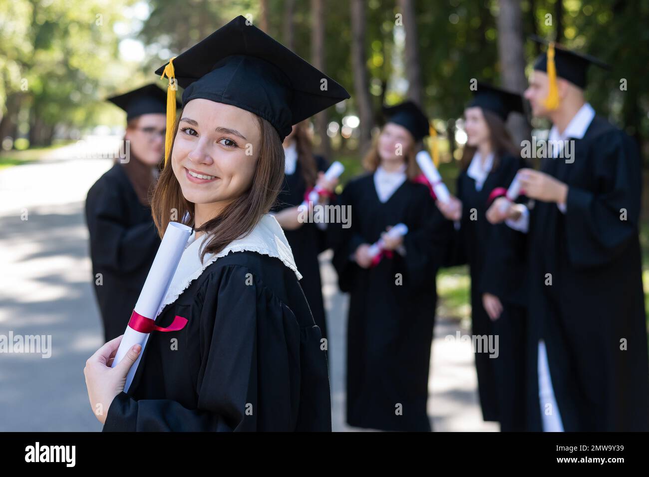 Group of happy students in graduation gowns outdoors. A young girl is ...