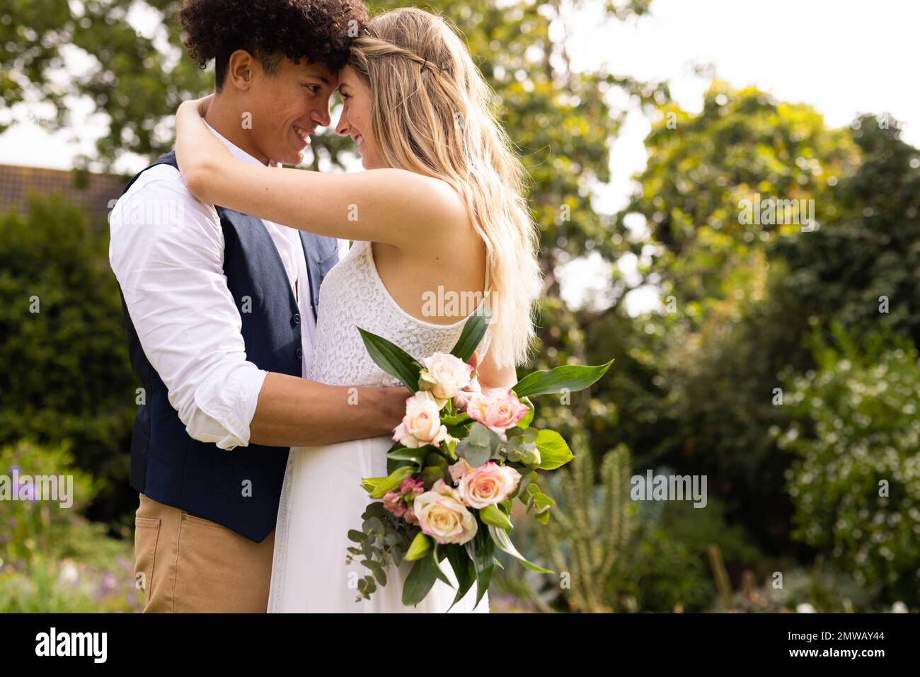 Happy diverse bride and groom with bouquet embracing at wedding in ...