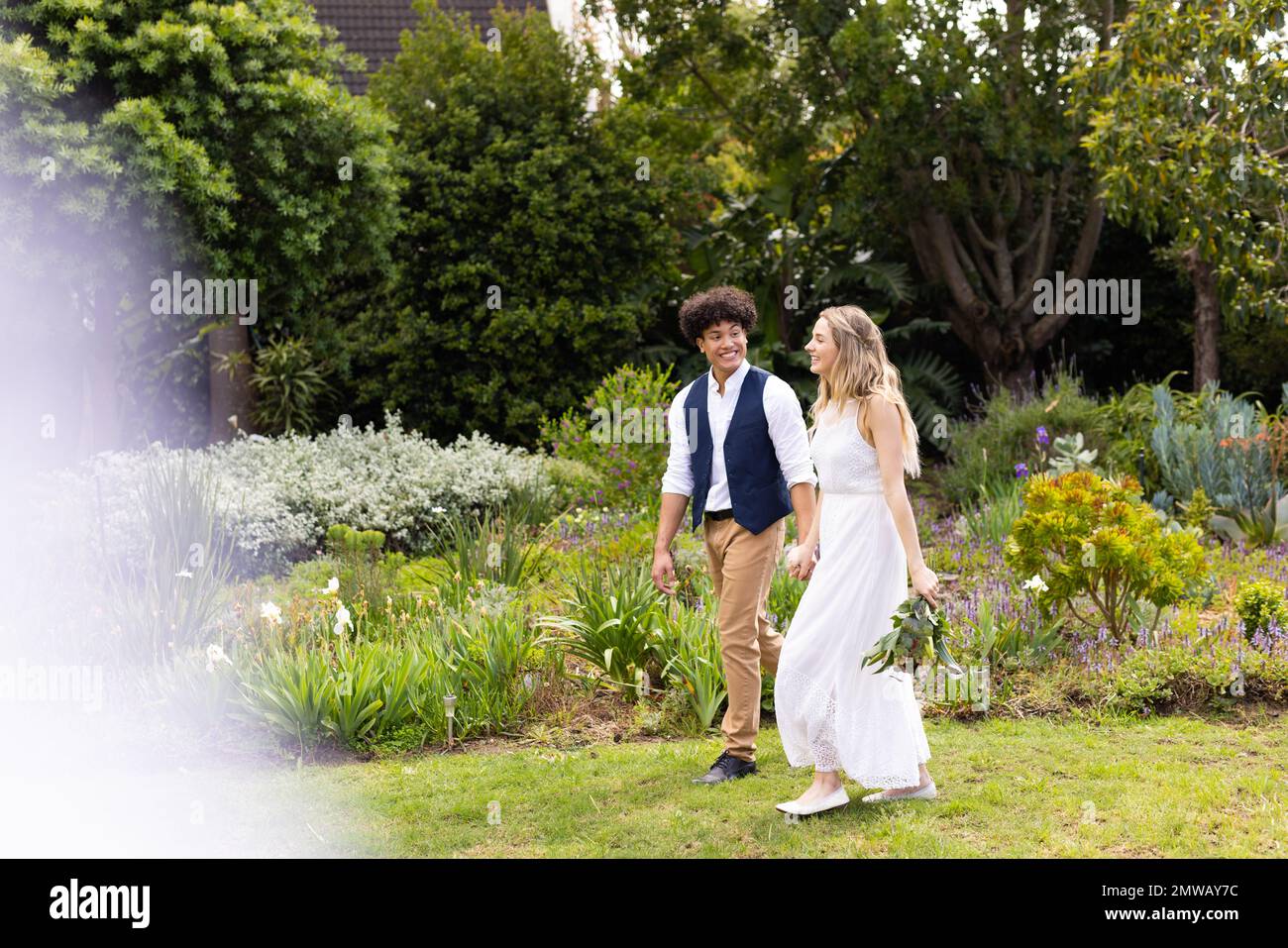 Happy diverse bride and groom holding hands, walking in garden at ...