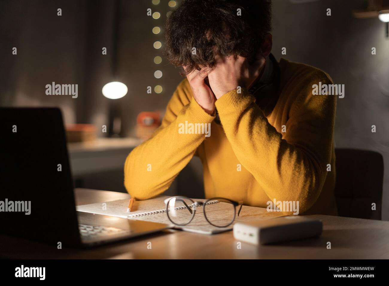 Tired and exhausted young man hides eyes with hands at laptop pc late ...