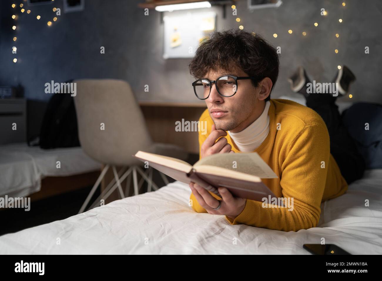 Teenage boy reading a book while laying on a bed in dormitory. Copy ...