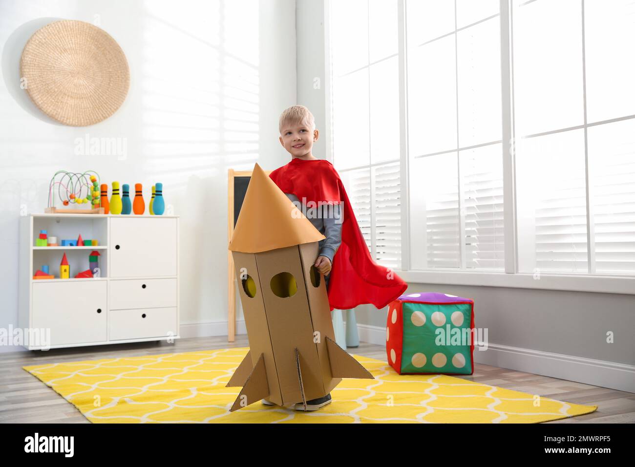 Little child in red cape playing with rocket made of cardboard box at ...