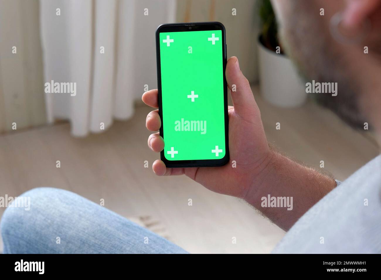 Young man sitting at home holding smartphone green mock-up screen in ...