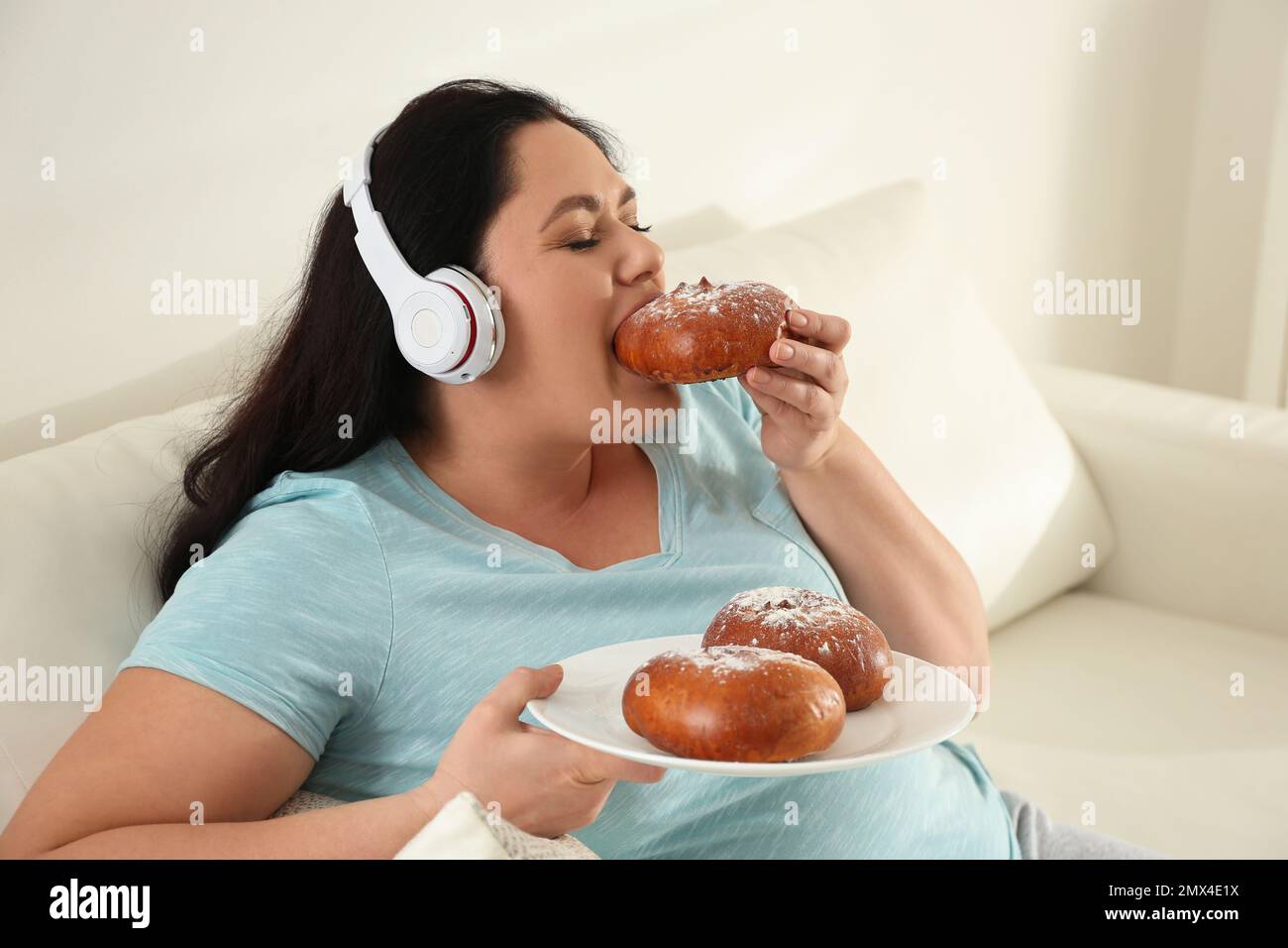 Lazy overweight woman with headphones eating bun at home Stock Photo ...
