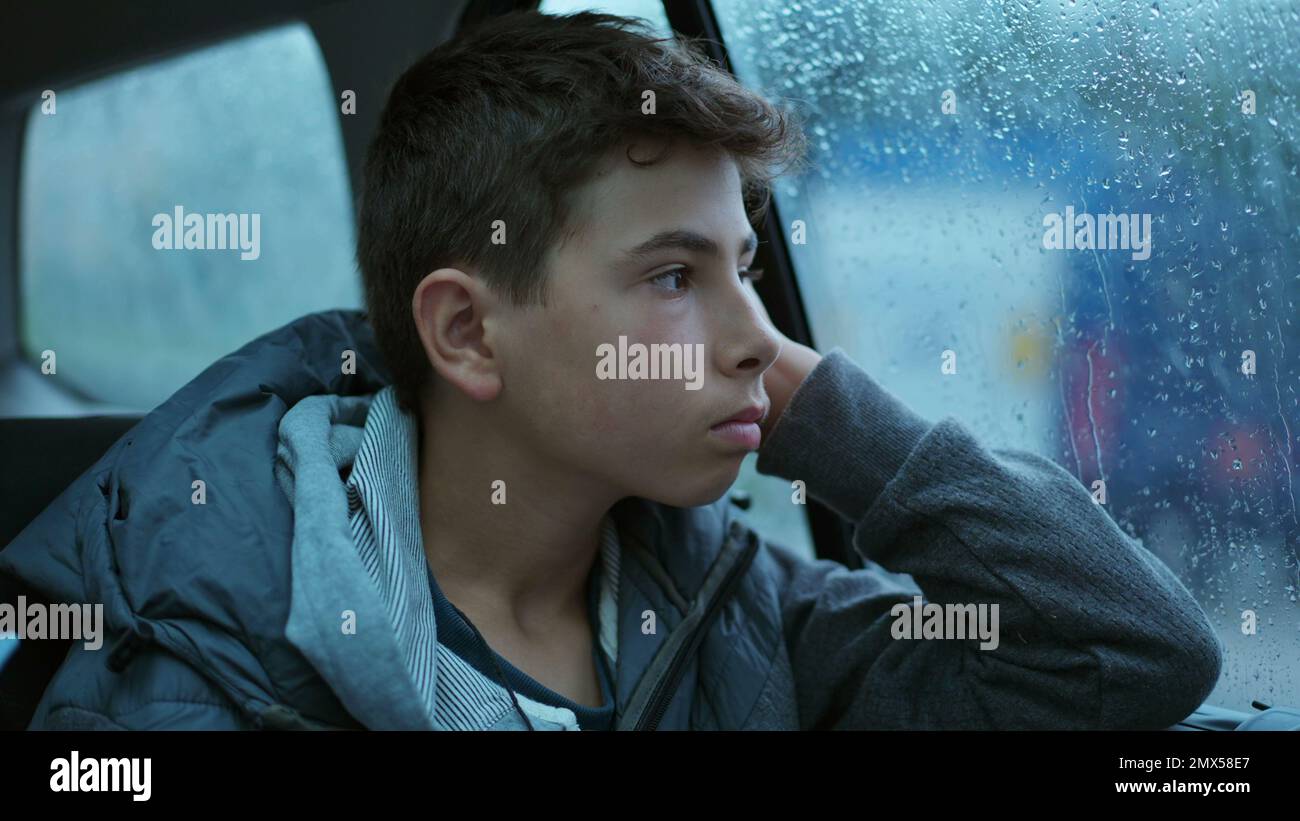 Thoughtful young boy seated in car backseat staring at glass window ...