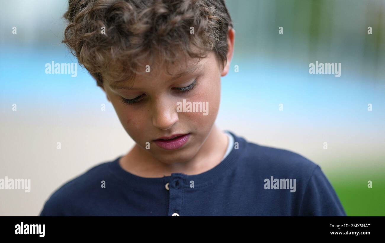 Portrait of a pensive young boy standing outside looking down in ...