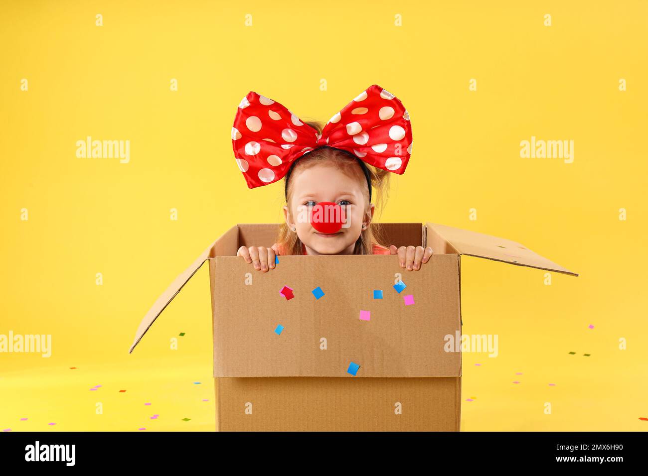 Little girl with large bow and clown nose in cardboard box on yellow ...