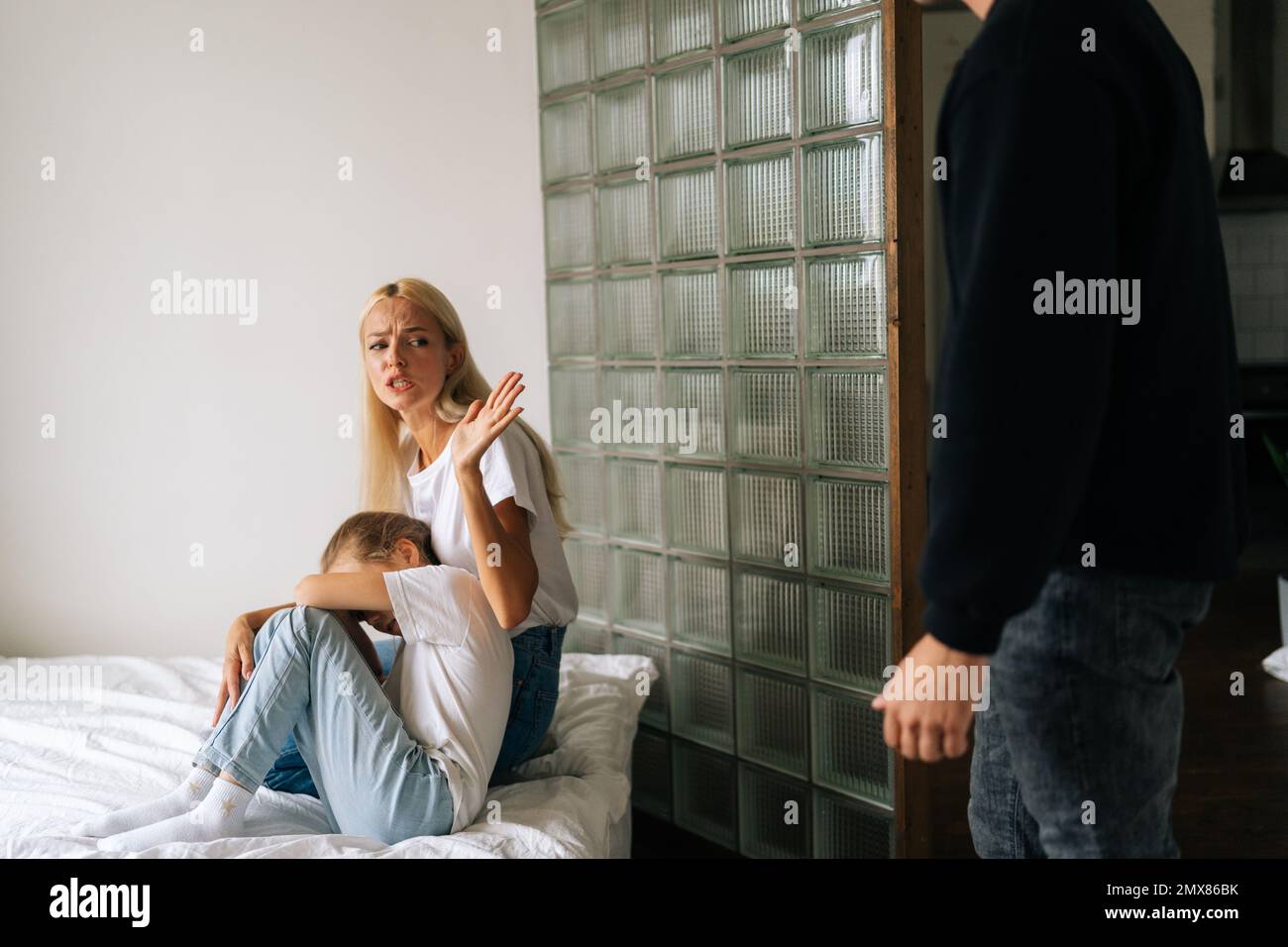 Portrait of scared mother and daughter hugging each other sitting on sofa while aggressive ...