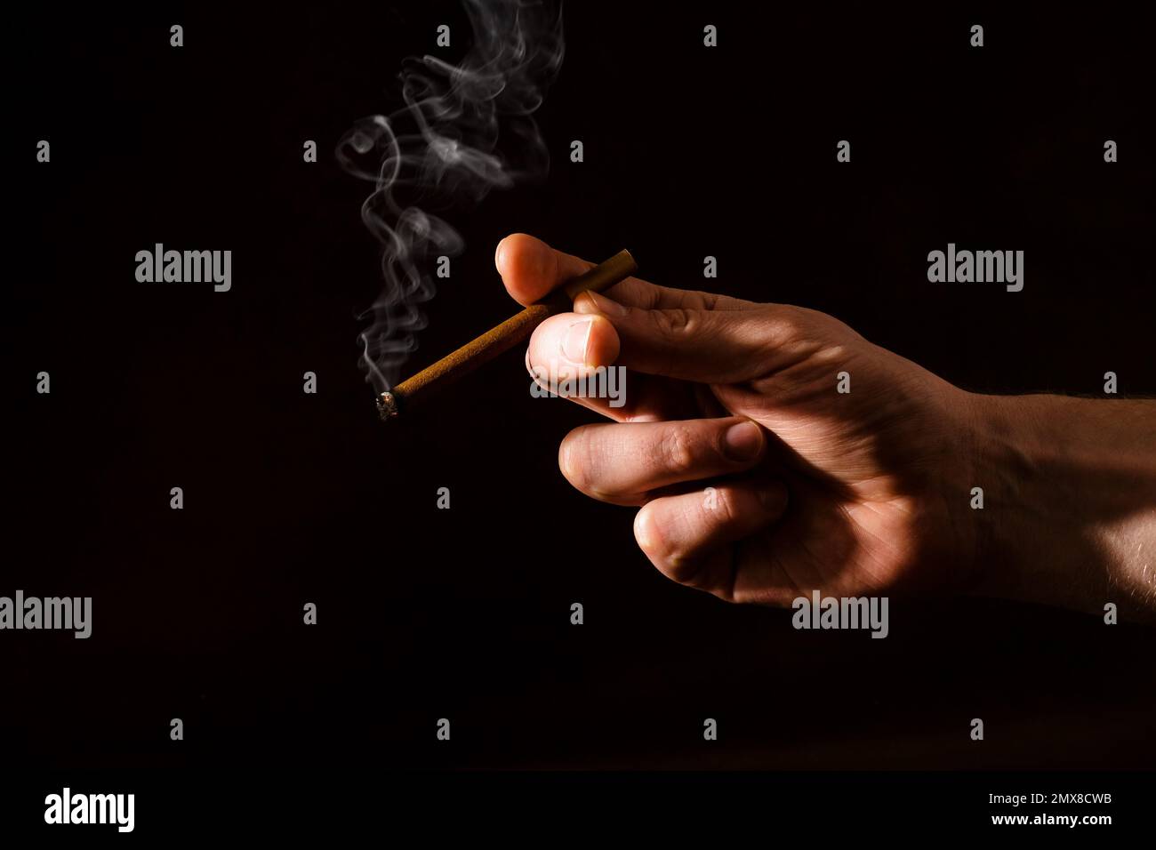 A man's hand holds a lit cigarette on a dark background. Hard light ...