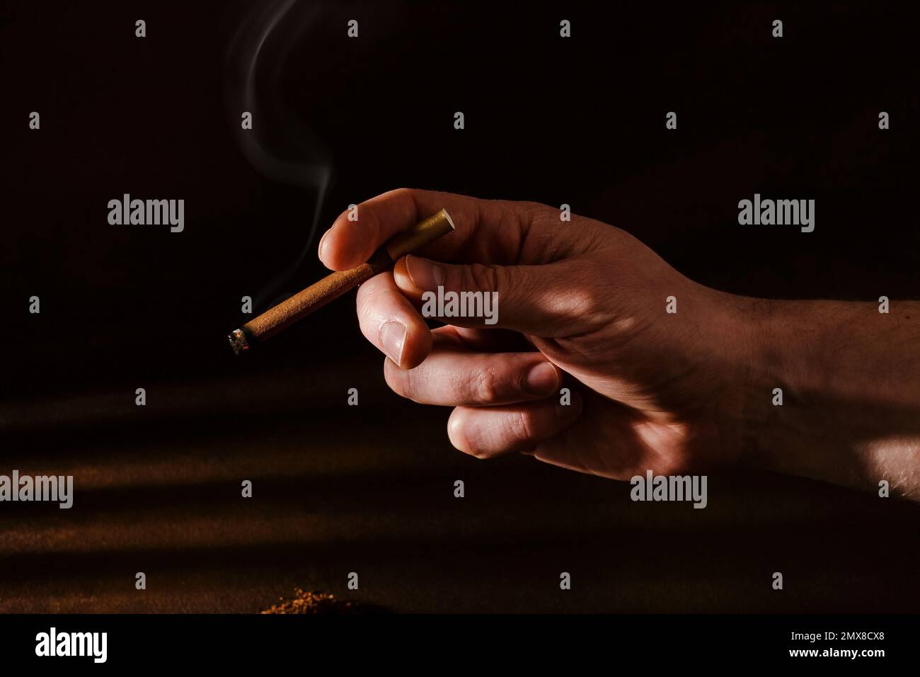 A man's hand holds a lit cigarette on a dark background. Hard light ...