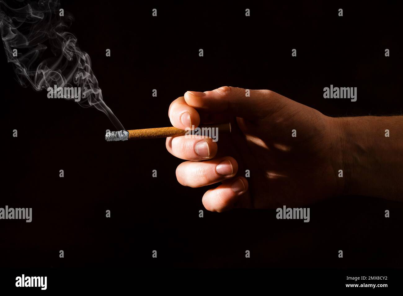 A man's hand holds a lit cigarette on a dark background. Hard light ...