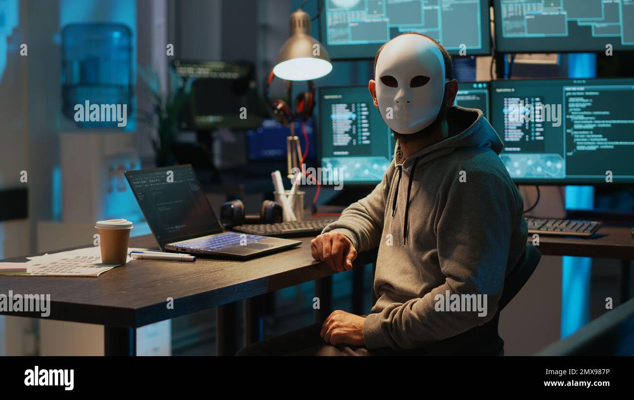 Masked scammer hacking computer network late at night, working on laptop and multiple monitors ...