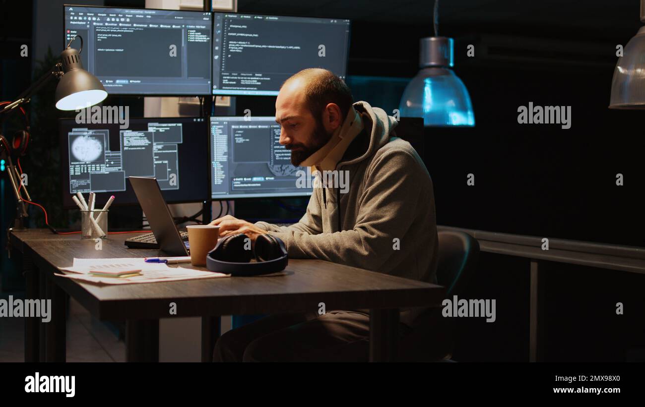 Software engineer wearing cervical collar at office, typing server code on multiple screens late ...