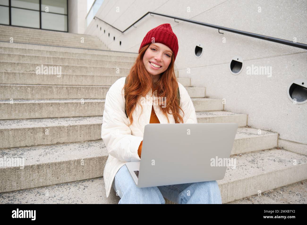 Smiling redhead girl, student sits on stairs outdoors and uses laptop ...