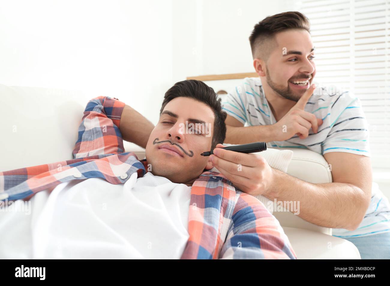 Young man drawing mustache on face of sleeping friend indoors. April ...