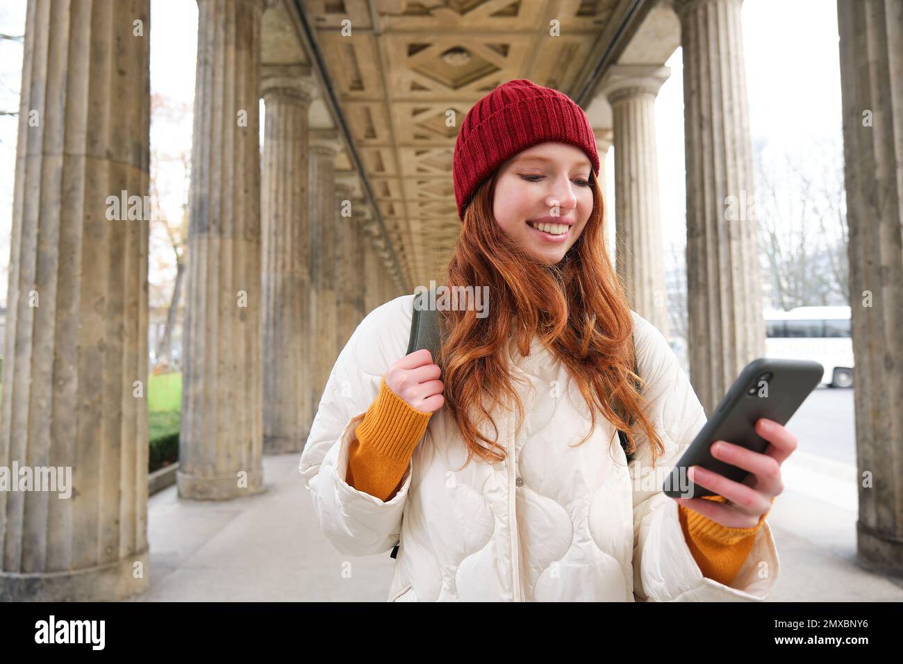 Mobile broadband and people. Smiling redhead 20s girl with backpack, uses smartphone on street ...