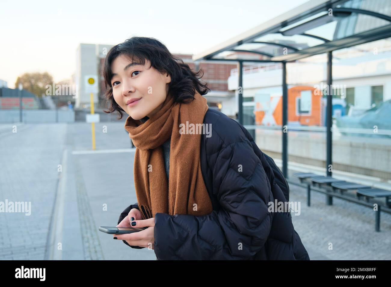 Cute smiling woman standing on a bus stop and looking at road, waiting ...