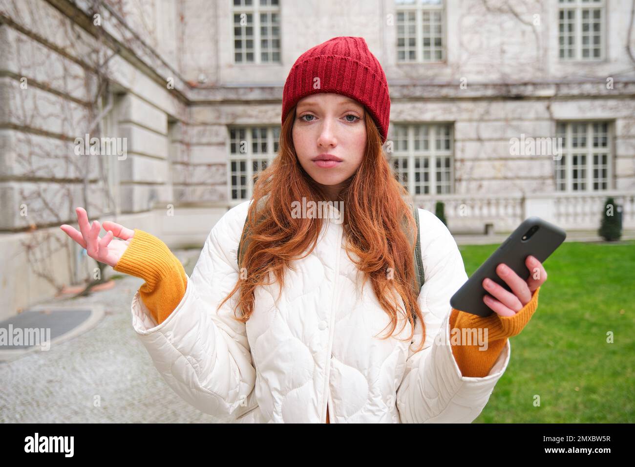 Portrait of confused redhead girl with smartphone, shrugs shoulders ...