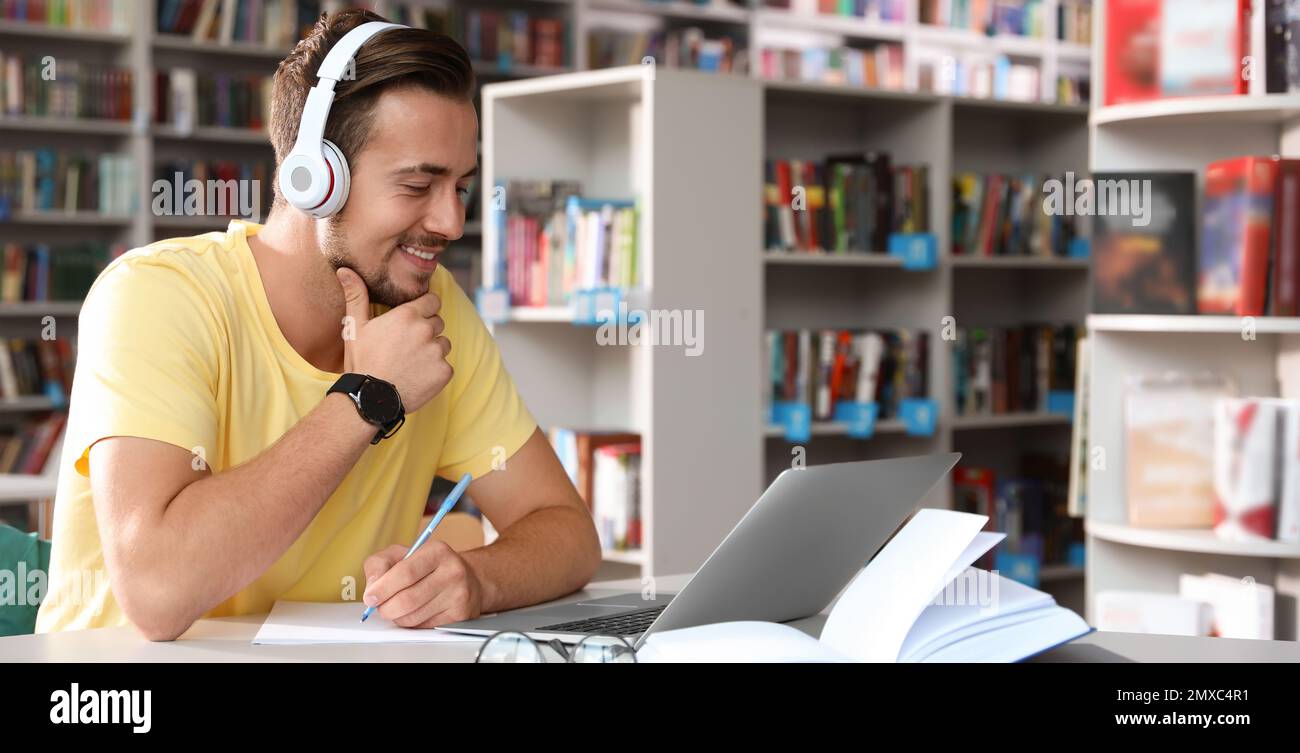 Young student wearing headphones at table in modern library, space for ...