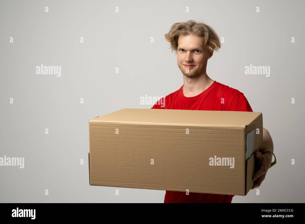 Smiling young delivery man in red t-shirt standing with big parcel post ...