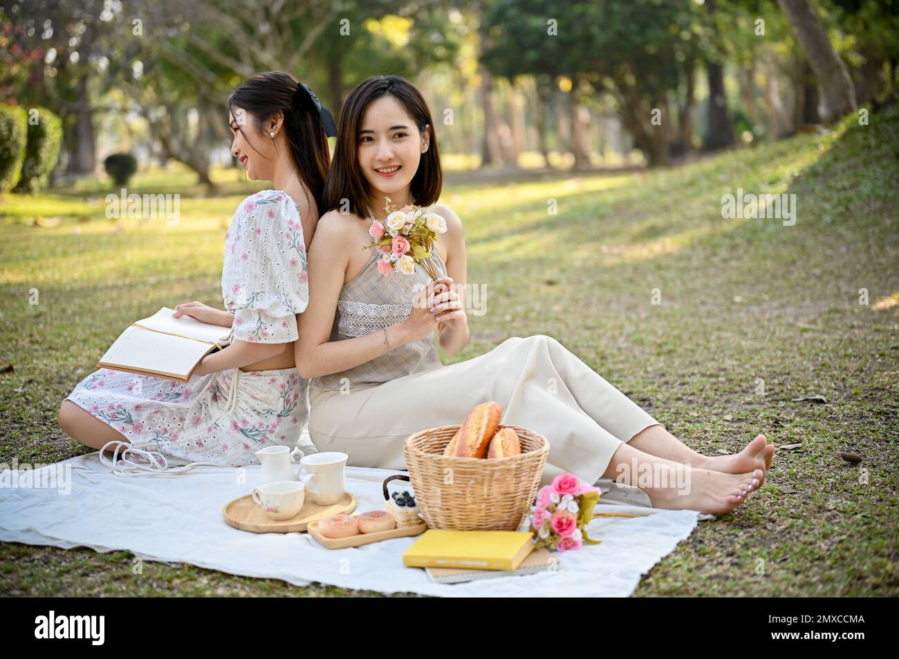 Two beautiful Asian women picnicking in the garden, sitting leaning on ...