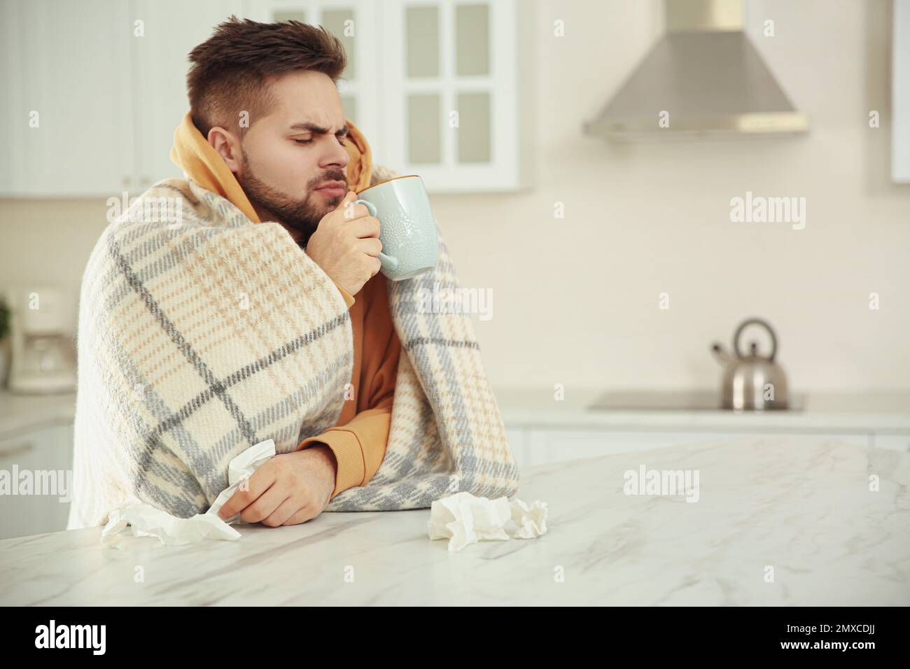 Sick young man with cup of hot drink and tissues in kitchen. Influenza ...