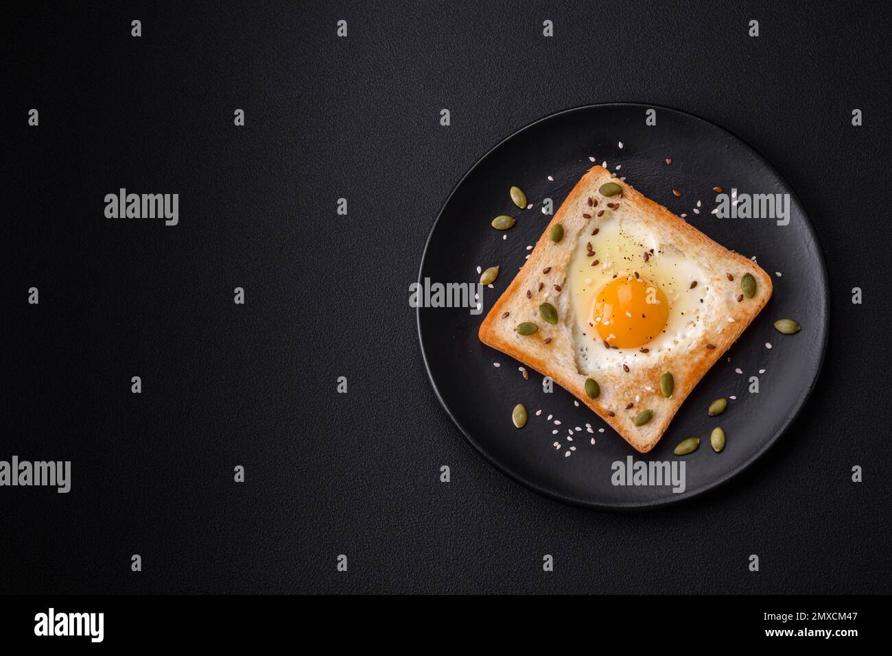 Heart shaped fried egg in bread toast with sesame seeds, flax seeds and pumpkin seeds on a black ...