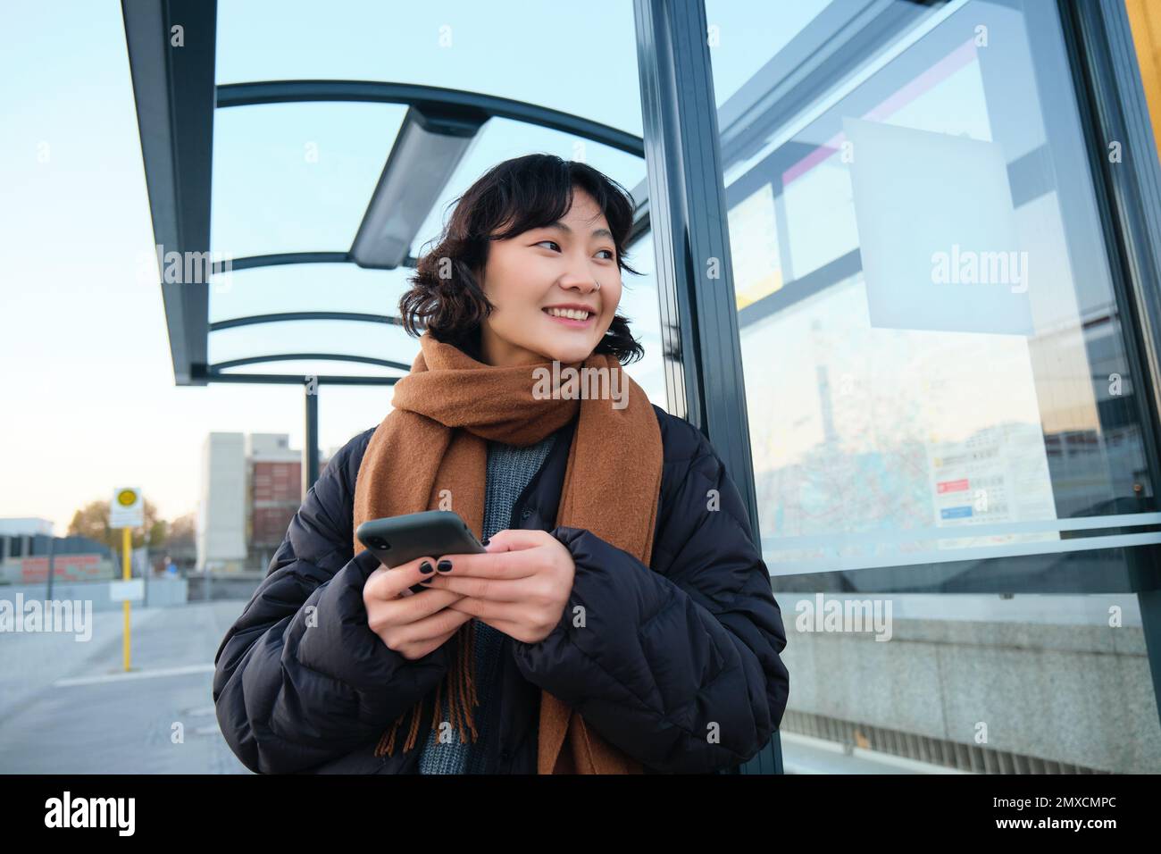 Cute smiling asian girl standing on bus stop, holding smartphone ...