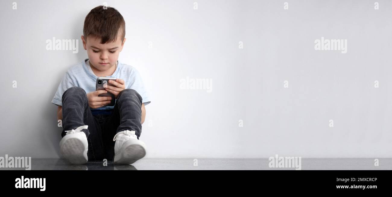 Sad little boy with mobile phone sitting near white wall, banner design ...