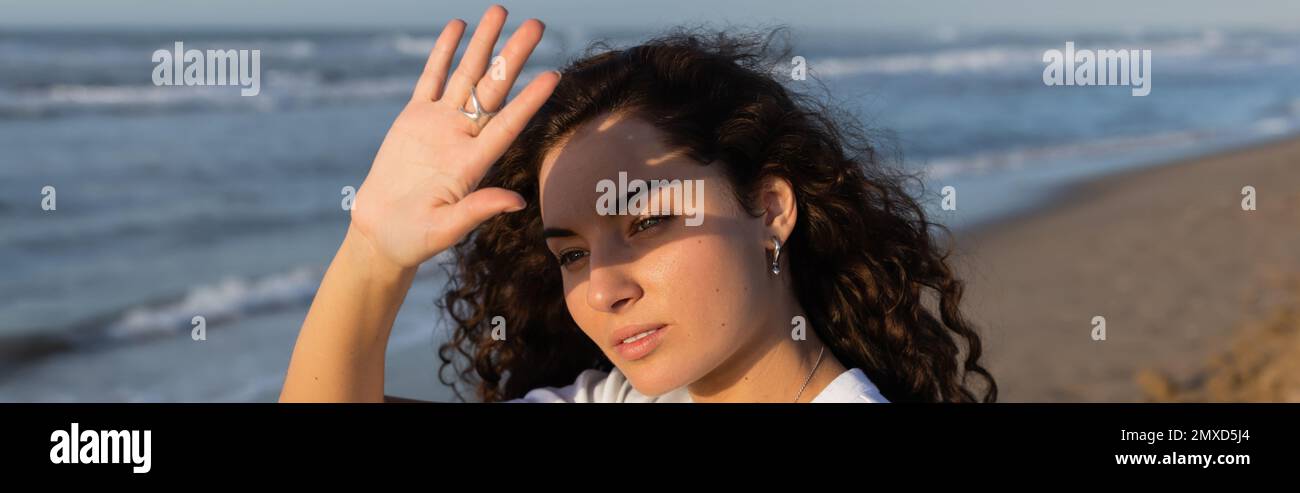portrait of curly woman hiding face from sun on beach near sea, banner ...