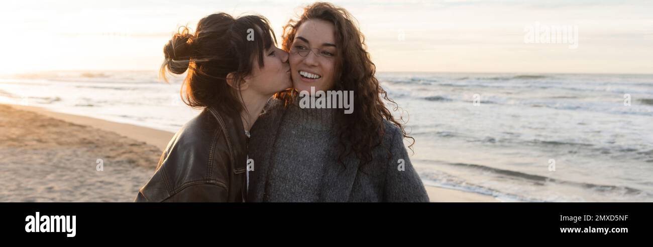 Young woman kissing cheeks of cheerful friend on beach in Spain, banner ...