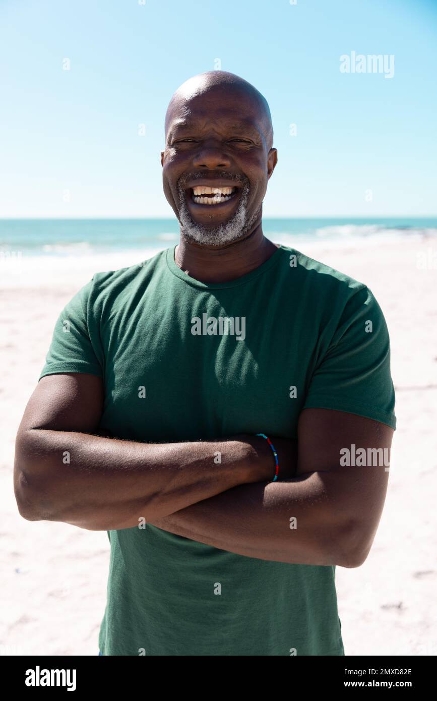 Portrait of bald african american senior man with arms crossed laughing at beach during summer ...