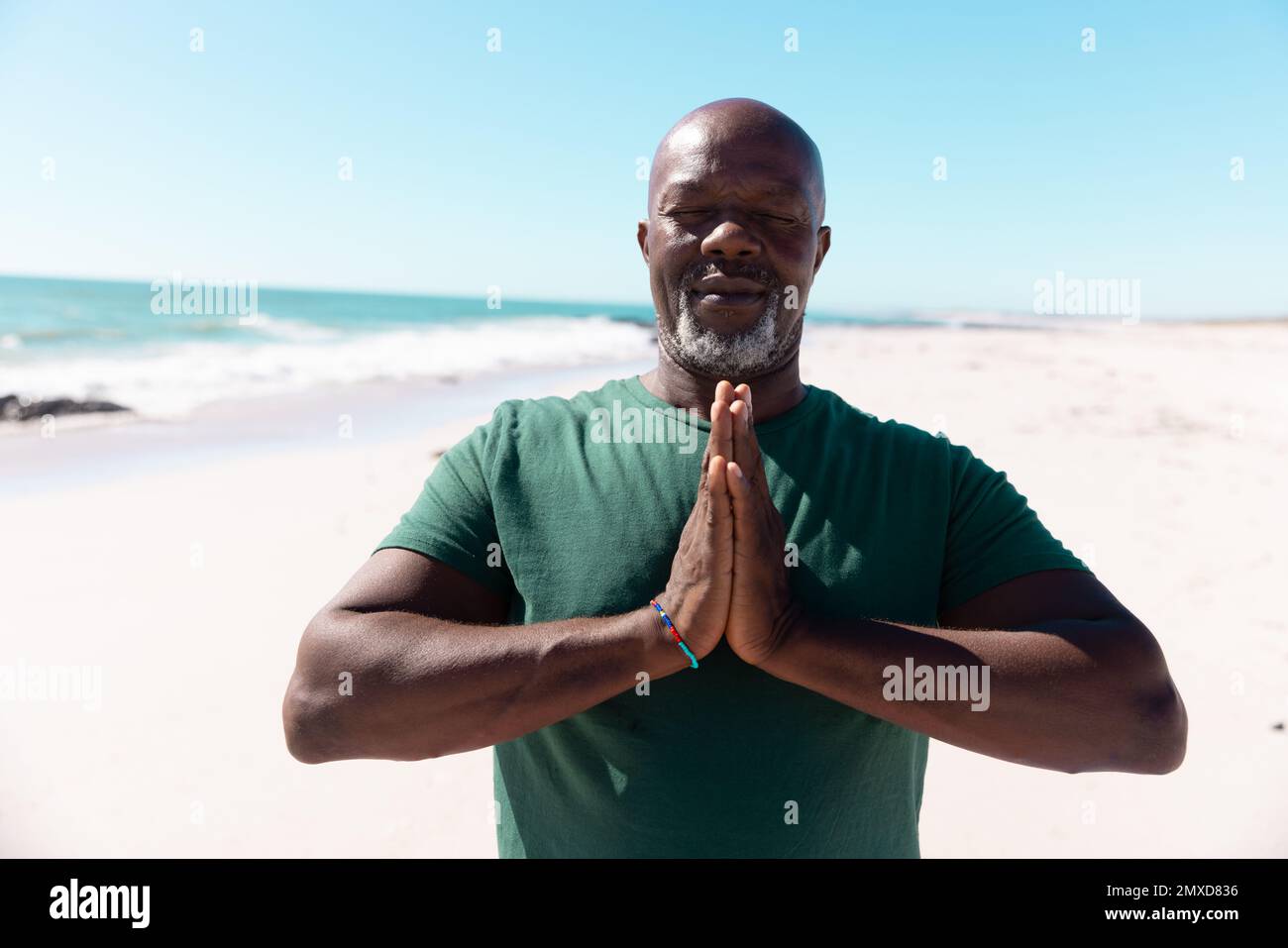 African american bald senior man with eyes closed meditating in prayer position at beach, copy ...