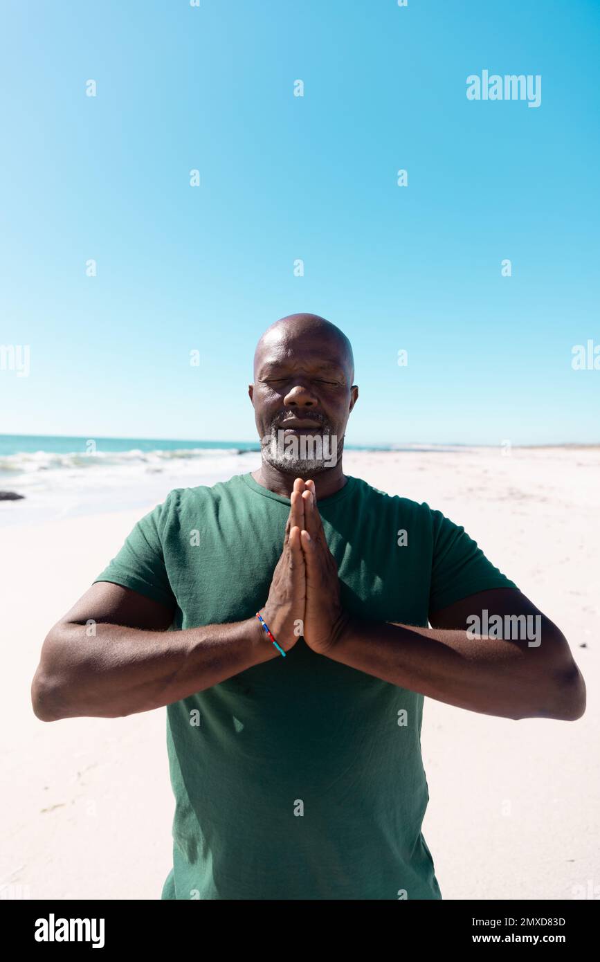 African american bald senior man with eyes closed meditating in prayer position at beach, copy ...