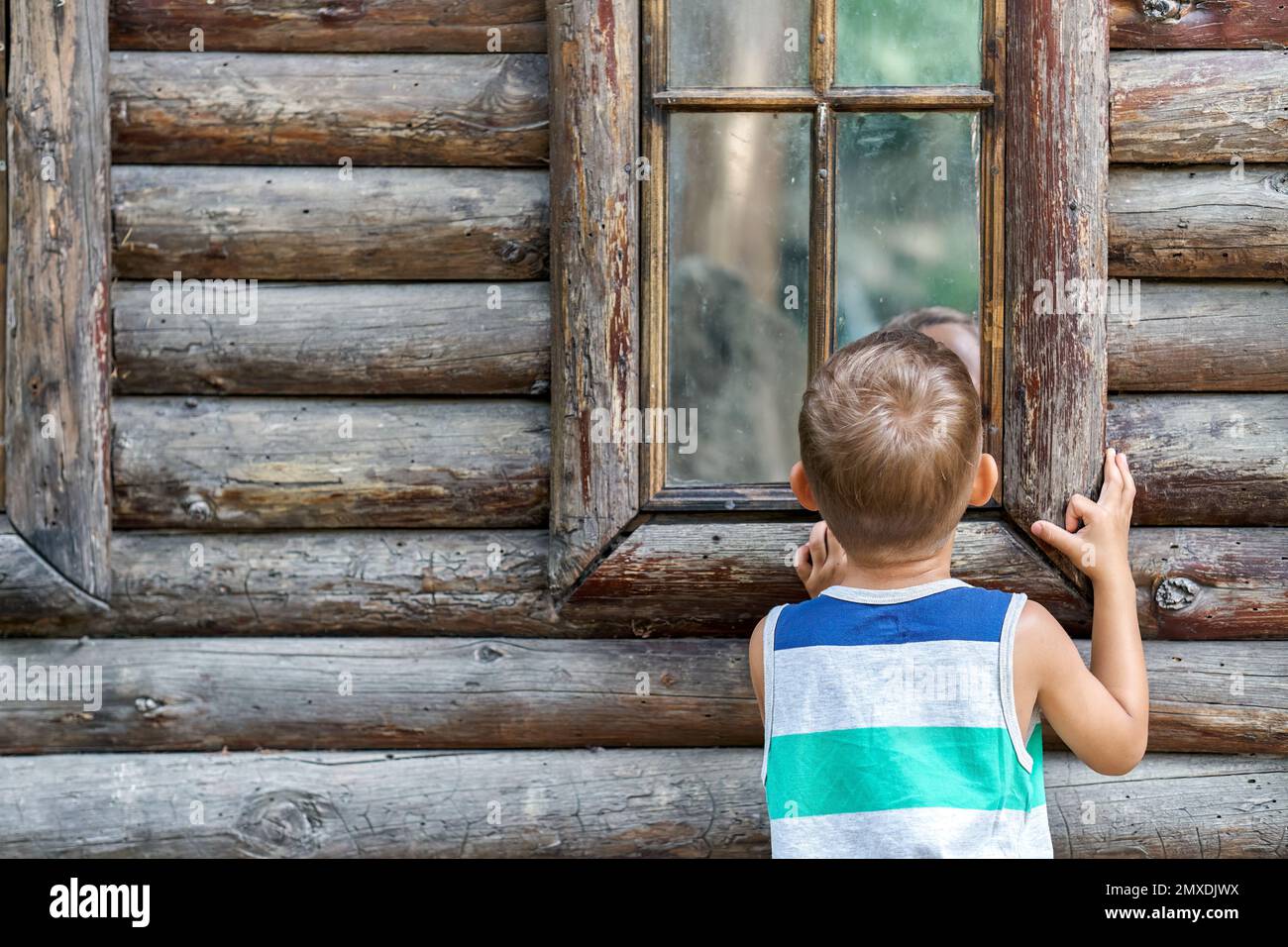 Excited preschooler boy looks into window of ancient log house. Curious ...