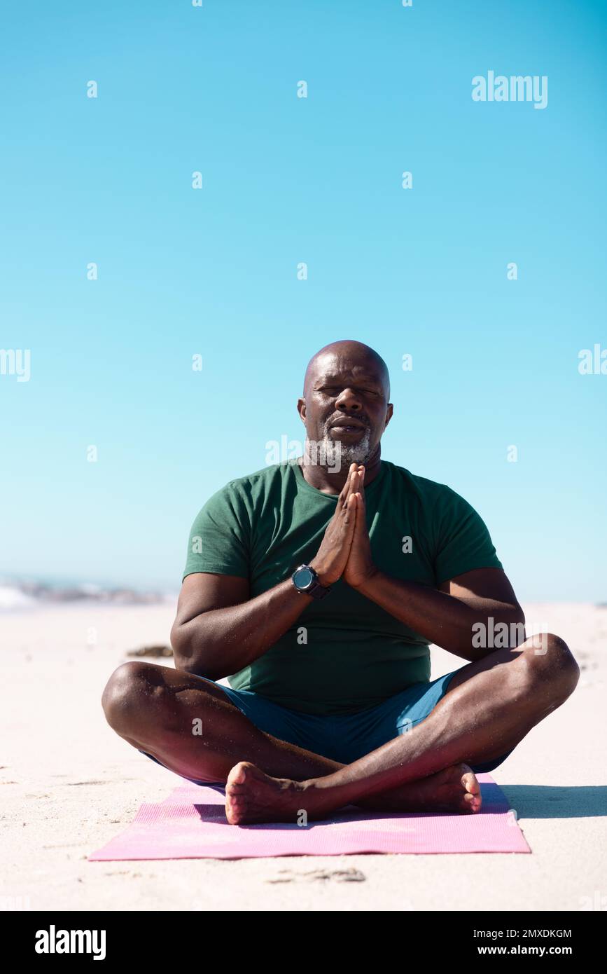 Bald african american senior man sitting cross legged and meditating at beach under clear blue ...