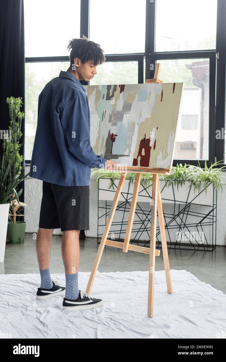 Side view of young african american artist holding painting in workshop ...