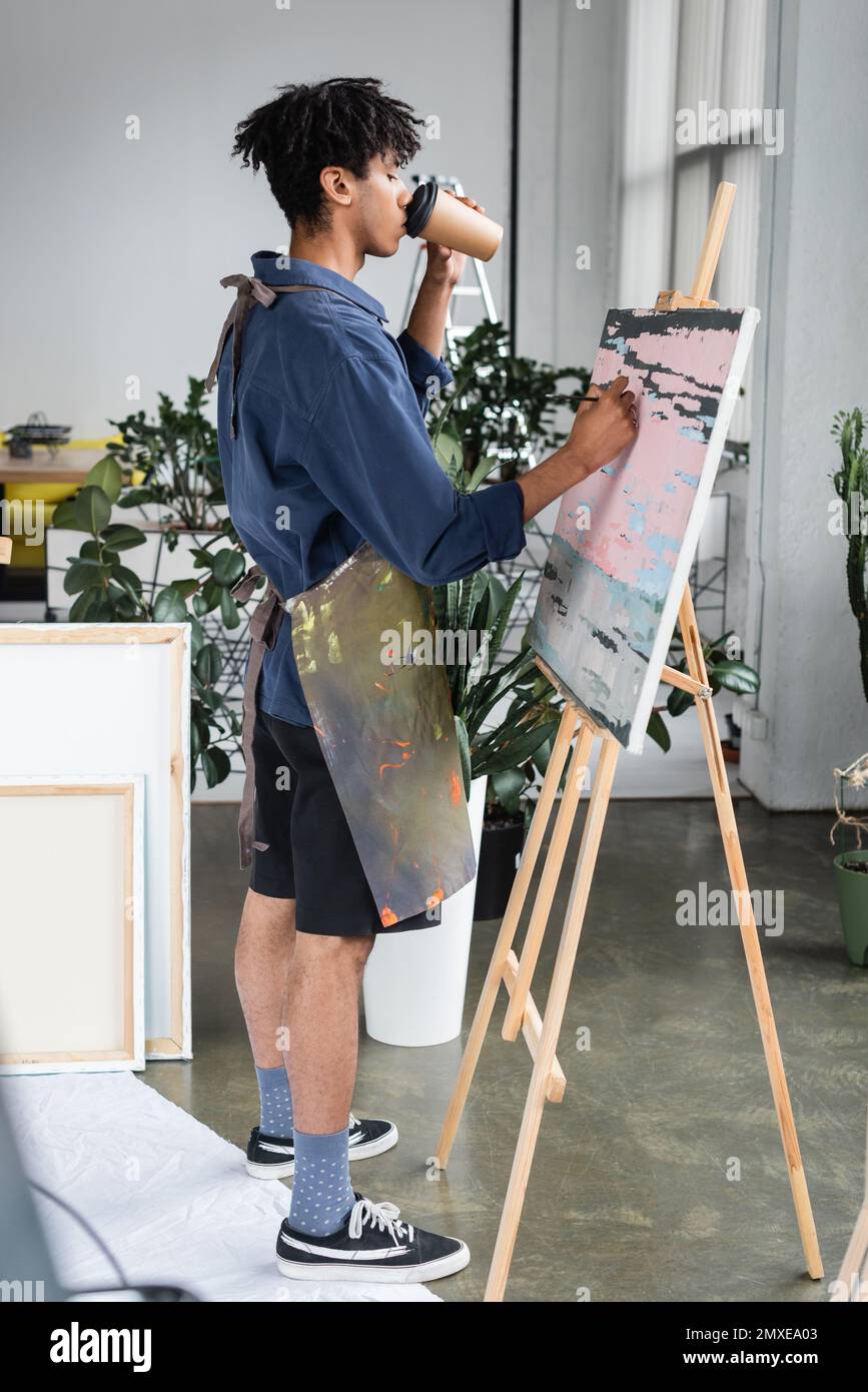 Side view of young african american artist drinking coffee and painting ...
