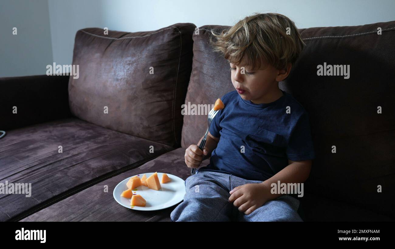 Cute boy eating healthy dessert fruit sitting on sofa at home child ...
