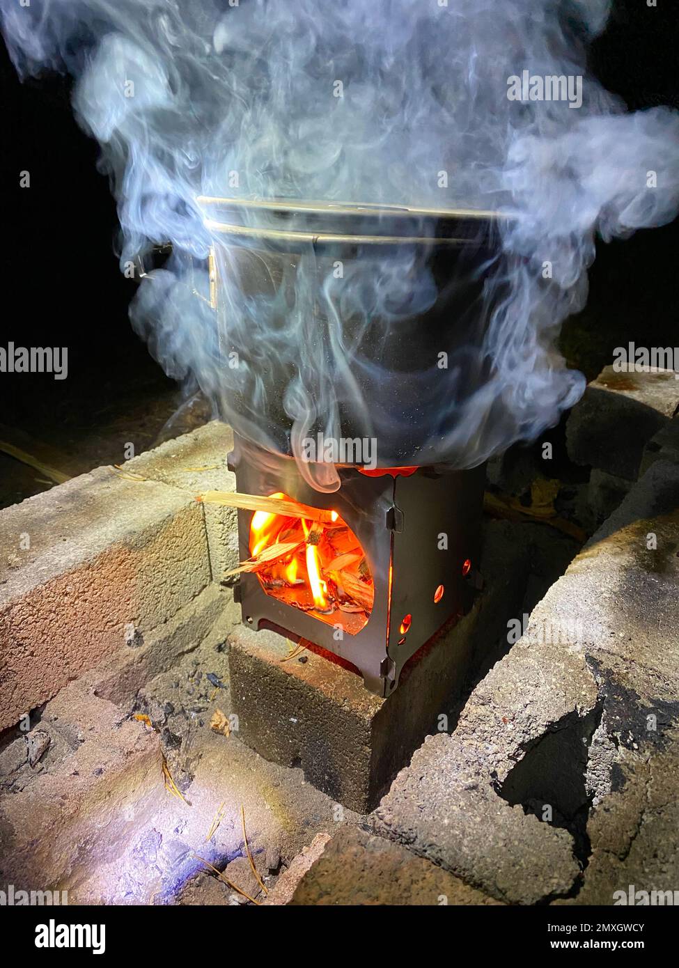 A wood chip burner burns at night from a pot with smoke on bricks ...