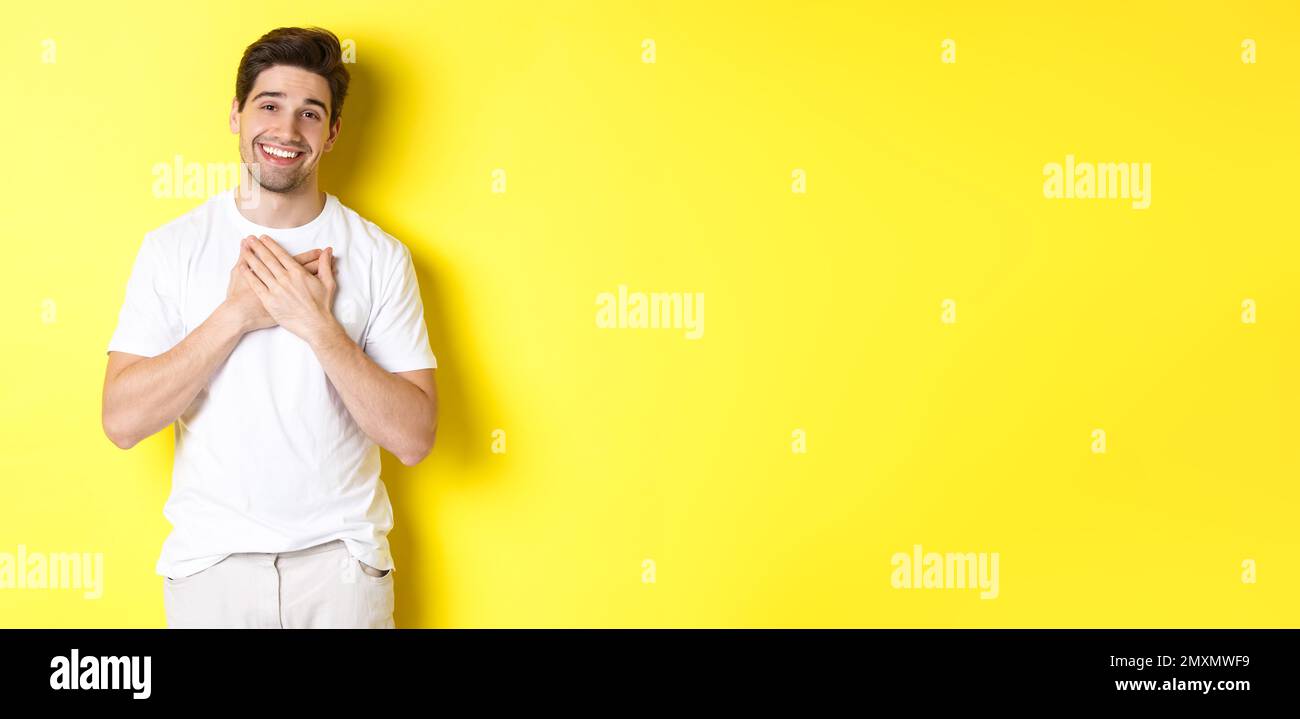 Image of grateful handsome guy in white t-shirt, holding hands on heart ...