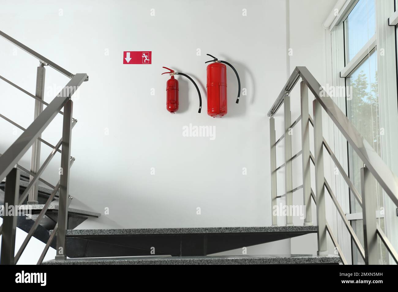 Fire extinguishers and emergency exit sign on white wall near staircase ...