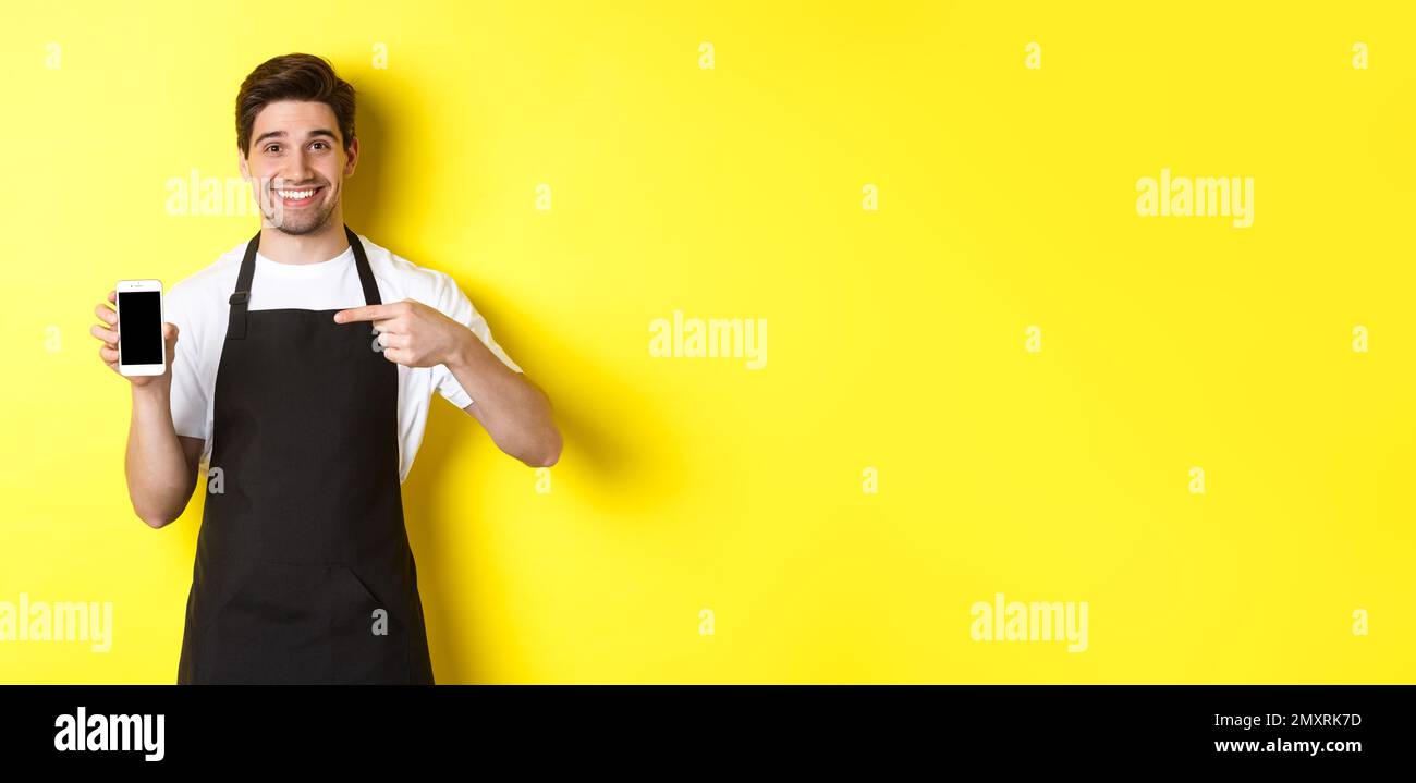 Handsome barista in black apron pointing finger at mobile screen ...